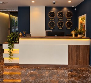 Bancada de recepção com relógios mostrando diferentes cidades, uma planta em vaso e um painel de madeira decorativo.