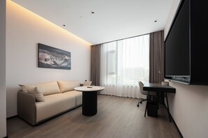 Quarto de hotel moderno com sofá, mesa redonda, escrivaninha, TV de tela plana e uma grande janela com cortinas.