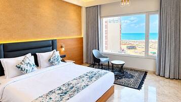 Quarto de hotel com cama, poltrona, mesinha, ventilador de teto e vista para a praia.