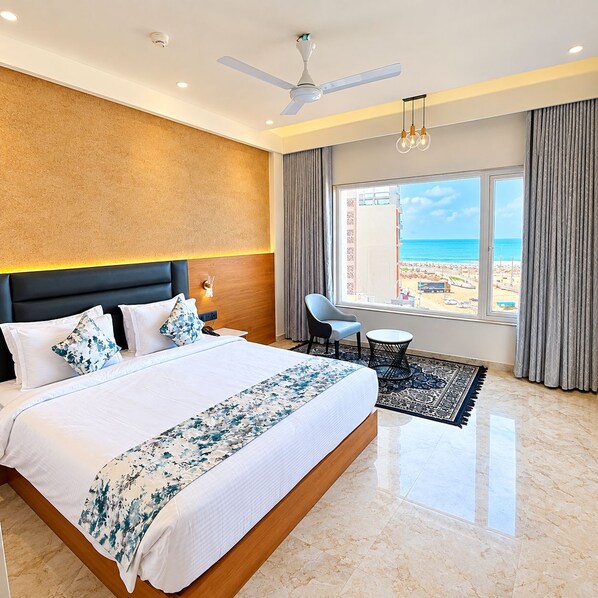 Quarto de hotel com cama, poltrona, mesinha, ventilador de teto e vista para a praia.
