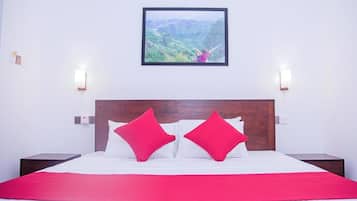 Quarto de hotel com uma cama, dois travesseiros vermelhos, um edredom branco e um quadro com uma paisagem na parede.