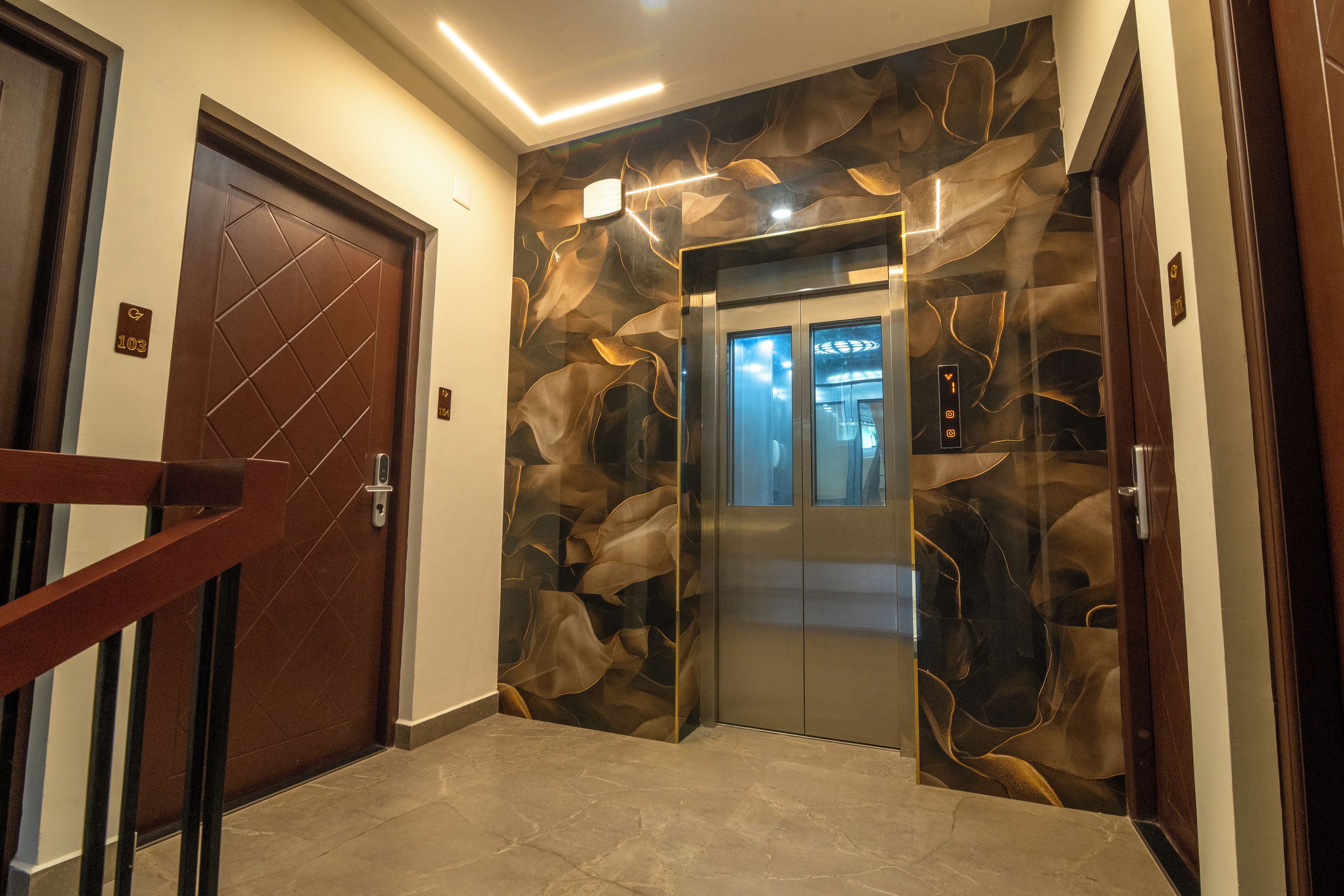 Entrada de hotel com porta de madeira, elevador de vidro e piso de mármore.
