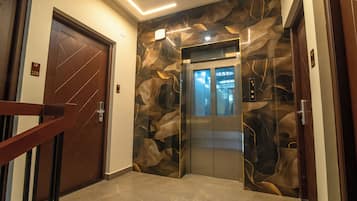 Entrada de hotel com porta de madeira, elevador de vidro e piso de mármore.