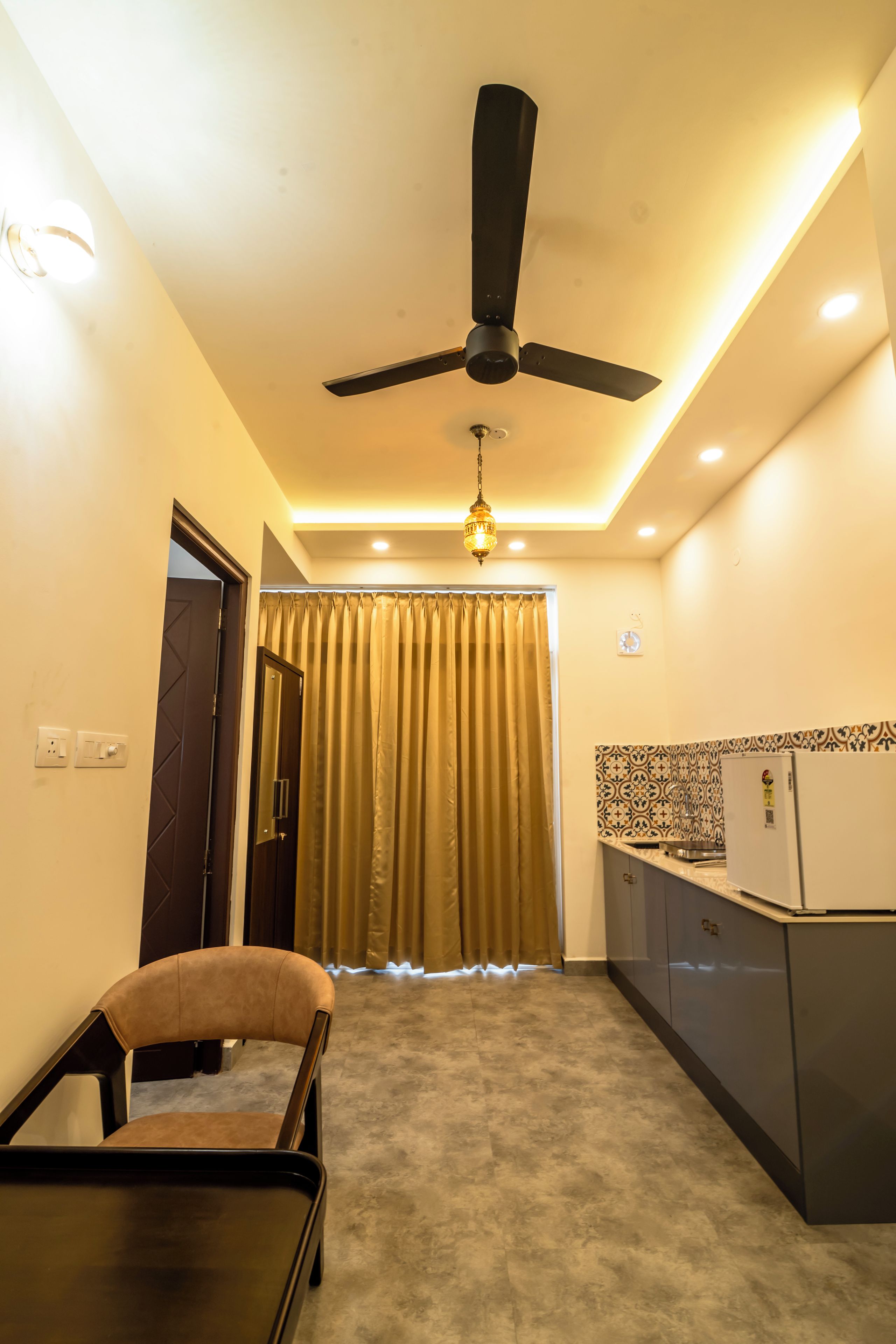 Uma sala moderna com ventilador de teto, mesa de jantar com uma cadeira, cozinha com geladeira e janela com cortinas.