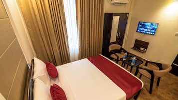 Quarto de hotel com cama, escrivaninha, cadeira, televisão e janela com cortinas.