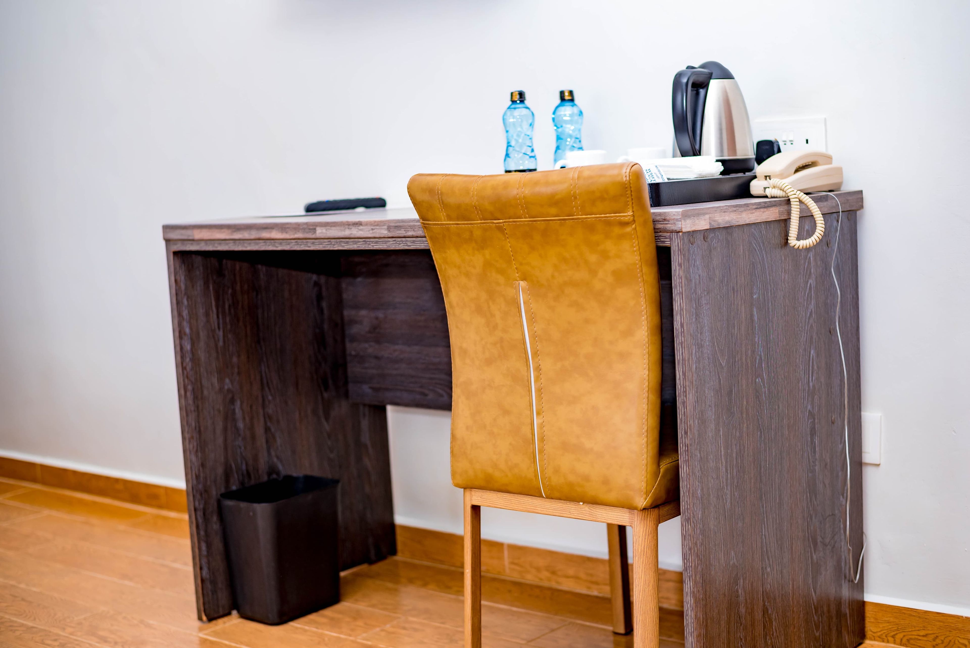 Uma mesa de madeira com cadeira de couro marrom, uma lixeira preta e um telefone sobre a mesa.