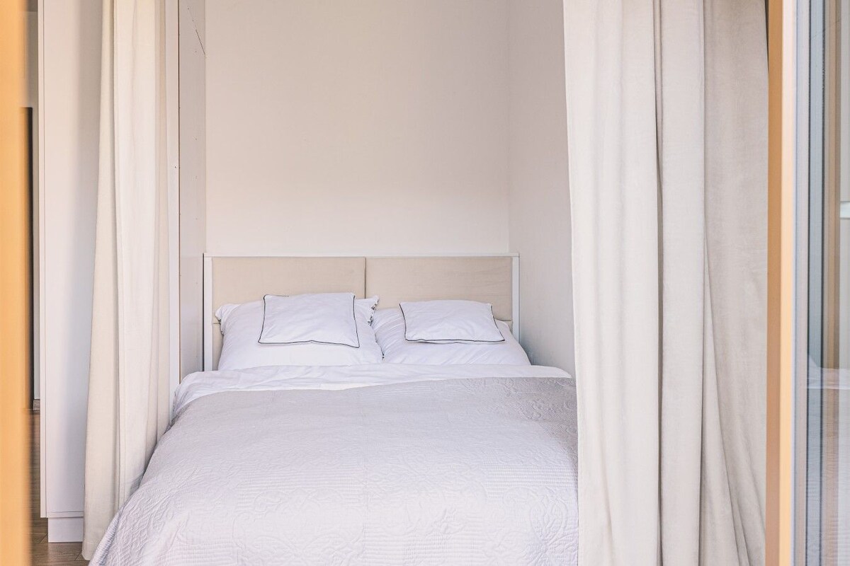 Cama com roupa de cama branca e cabeceira, cortinas semi-abertas, e parede lisa.
