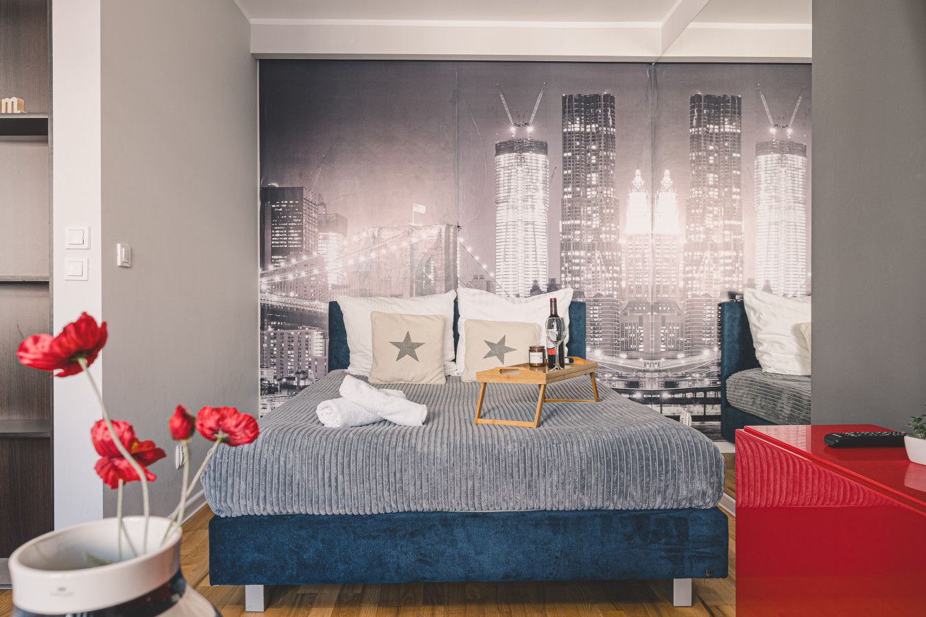 Quarto com mural da paisagem urbana, uma cama com almofadas, mesa de cabeceira com garrafa e pote, e mesa lateral vermelha.