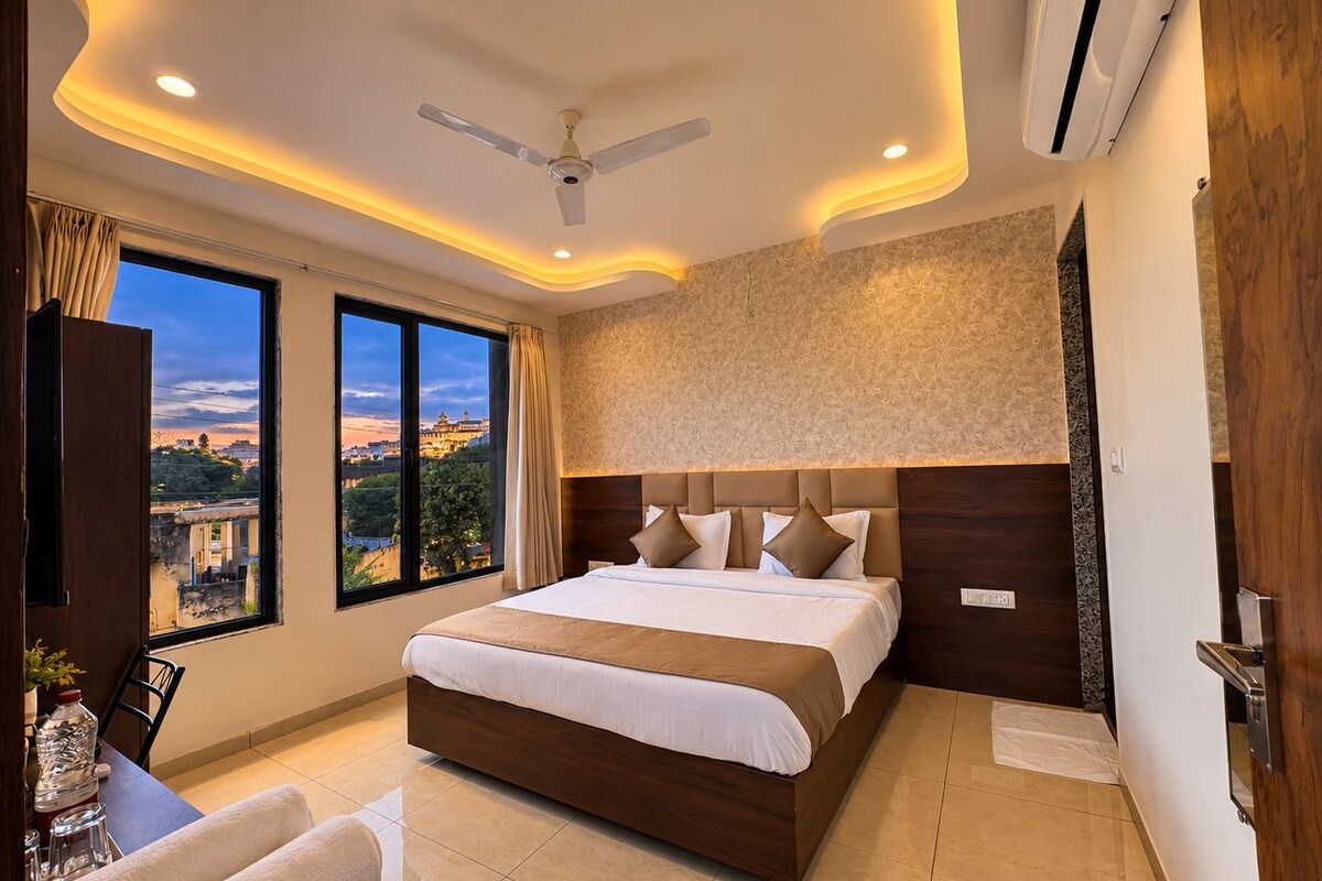 Quarto de hotel com uma cama grande, ventilador de teto, ar condicionado, vista para a cidade e área de jantar com mesa e cadeiras.