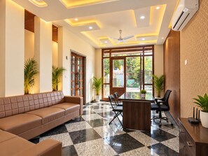 Um hall de hotel com piso xadrez, sofá de couro, balcão de recepção de madeira e plantas em vasos.