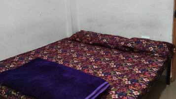 Cama de solteiro com colcha florida e manta roxa em quarto com paredes brancas e tomada visível.
