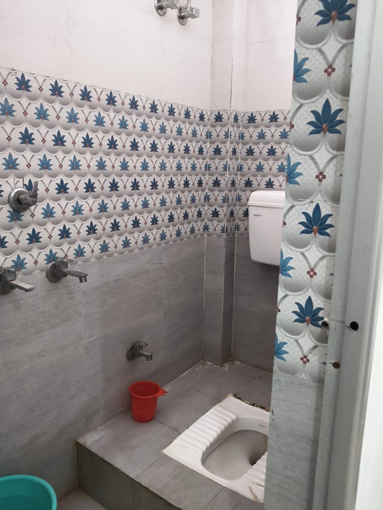 Um banheiro pequeno com vaso sanitário, um balde vermelho e cortina de chuveiro com padrão de folhas.