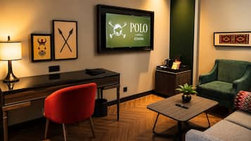 Quarto de hotel com escrivaninha, uma cadeira vermelha, uma poltrona verde, uma mesinha com uma planta e uma televisão exibindo 'POLO HOTEL KOHIMA'.
