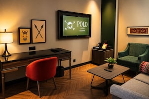 Quarto de hotel com escrivaninha, uma cadeira vermelha, uma poltrona verde, uma mesinha com uma planta e uma televisão exibindo 'POLO HOTEL KOHIMA'.