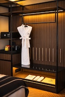 Quarto de hotel com armário contendo roupão branco, máquina de café e minibarra.