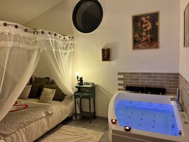 Quarto com cama, mesa de cabeceira com luminária, pintura e banheira com velas.