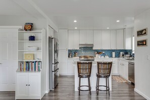 Cozinha moderna com armários brancos, forno embutido, geladeira de aço inoxidável e backsplash com azulejos azuis. Há dois banquetas de madeira na bancada.