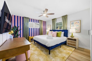Quarto de hotel com cama, mesas de cabeceira, escrivaninha, ventoinha no teto e cortinas listradas.