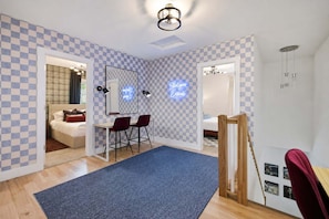 Quarto de hotel com padrão quadriculado na parede, uma cama, uma escrivaninha com duas cadeiras, um tapete azul e uma escada.