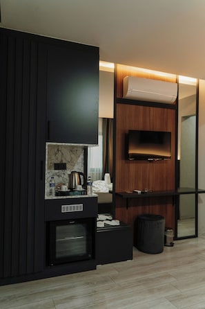 Quarto de hotel moderno com televisão de tela plana, cozinha compacta com cafeteira e lixeira.