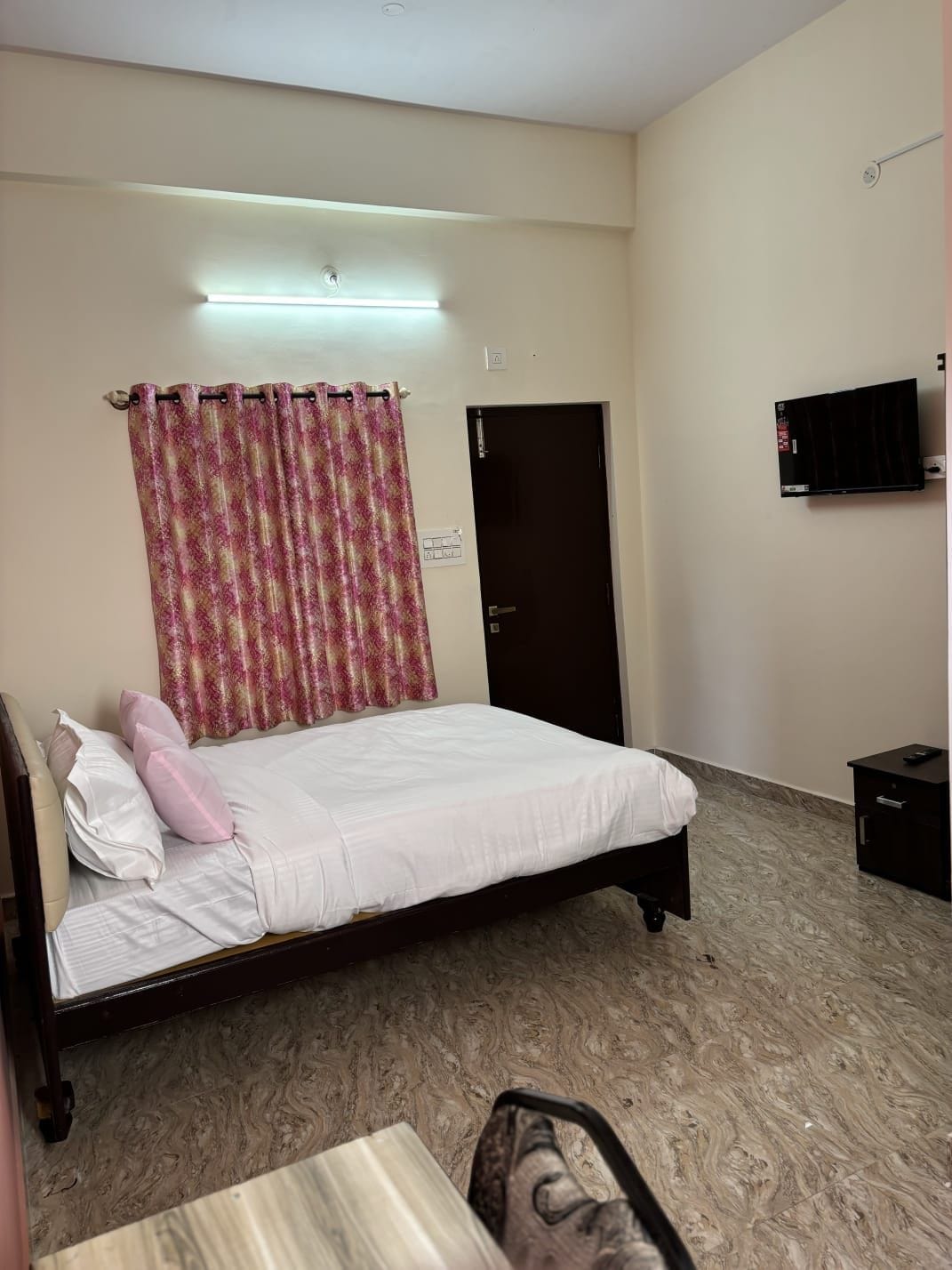 Quarto com cama, televisão na parede, uma porta e uma mesinha.
