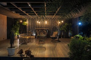 Uma área de pátio ao ar livre com piso de madeira, mesas e cadeiras dispostas, e uma parede decorativa com padrão de tijolos.