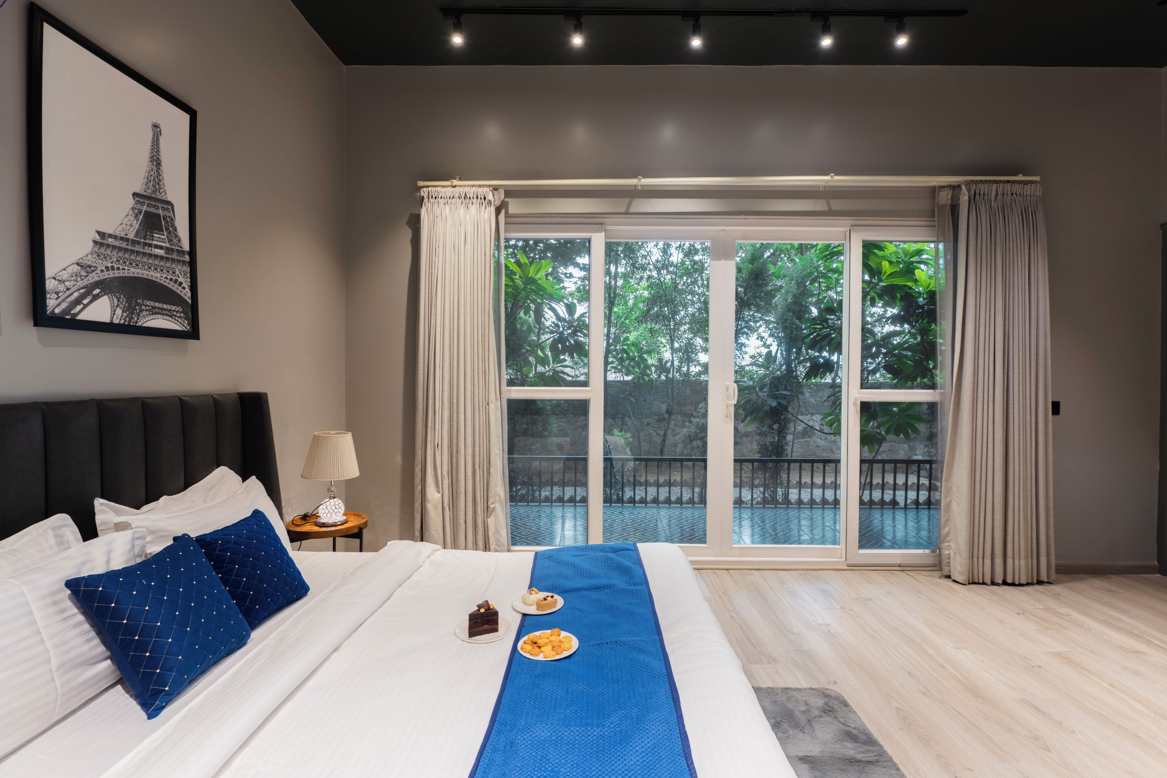 Quarto de hotel com uma cama grande, uma mesa de cabeceira, uma luminária, um quadro com a Torre Eiffel e vista para uma piscina e área verde através da janela.