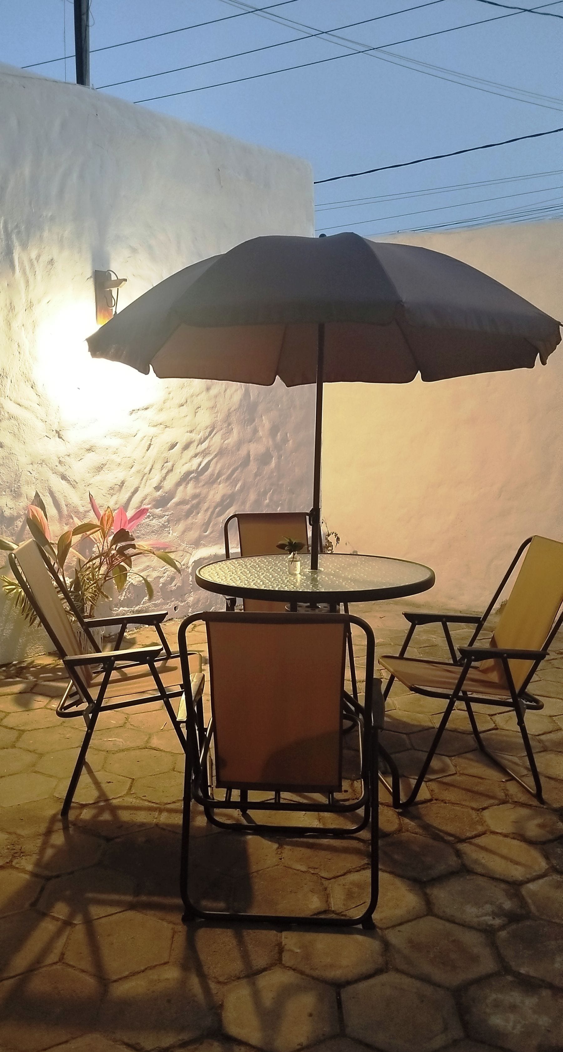 Um pátio ao ar livre com uma mesa redonda e quatro cadeiras, um guarda-sol e um vaso de planta.