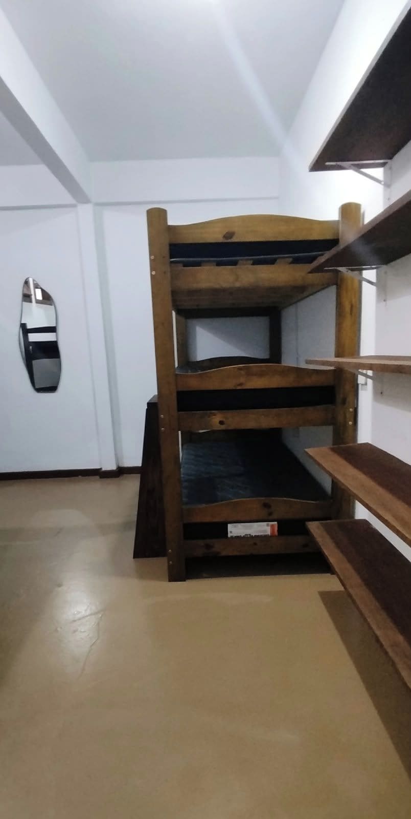 Uma cama de beliche com estrutura de madeira e grades de metal, uma parede branca com uma porta e um chão com uma costura visível.