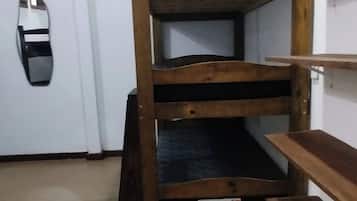 Uma cama de beliche com estrutura de madeira e grades de metal, uma parede branca com uma porta e um chão com uma costura visível.