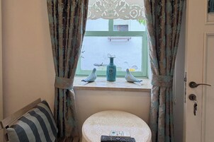 Um quarto com janela, cortinas, um sofá, um banquinho pequeno e um vaso com objetos decorativos no parapeito.