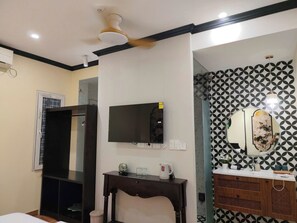 Quarto de hotel com cama, televisão na parede, mesinha com chaleira e espelho com moldura decorativa.