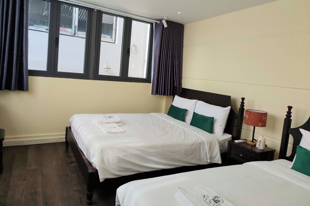 Quarto de hotel com duas camas, uma mesa de cabeceira com luminária e janela com cortinas.