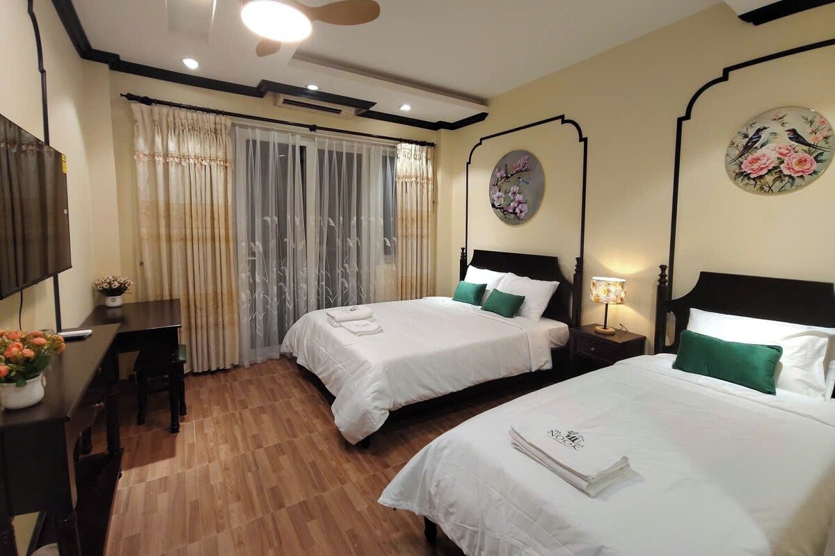 Quarto de hotel com duas camas, uma escrivaninha, uma cadeira, ventoinha no teto e janela com cortinas.