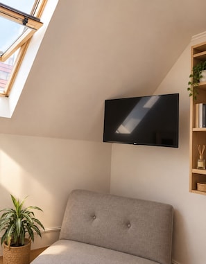 Quarto com televisão de tela plana na parede, sofá cinza, planta em vaso e estante com livros e um objeto decorativo.