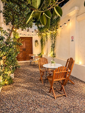 Pátio externo com mesa e cadeiras de madeira, cercado por pedras e vegetação.