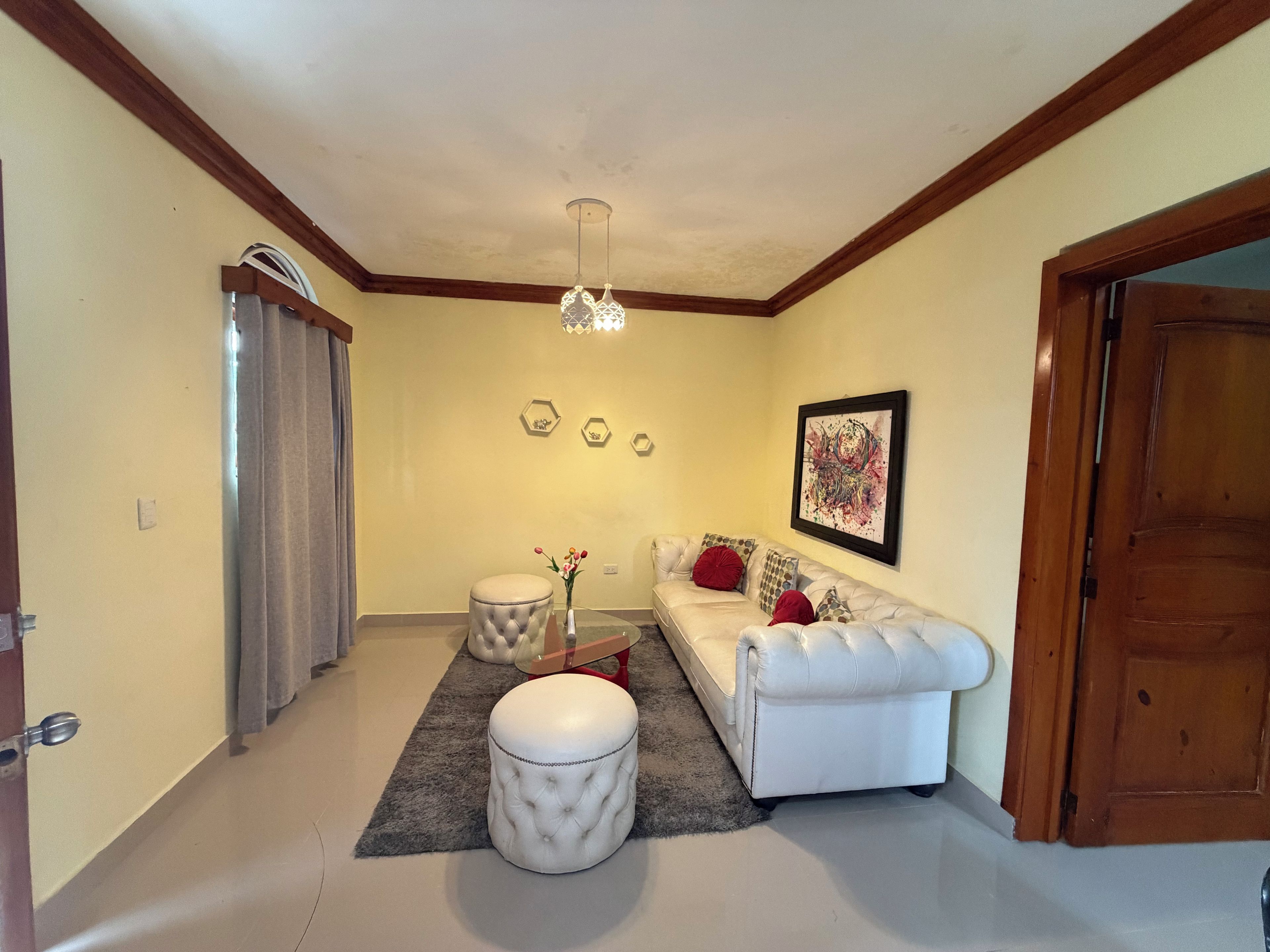 Sala de estar com sofá branco, um banquinho e uma mesa de centro. Há uma porta à direita e uma janela à esquerda.