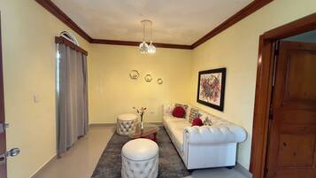 Sala de estar com sofá branco, um banquinho e uma mesa de centro. Há uma porta à direita e uma janela à esquerda.