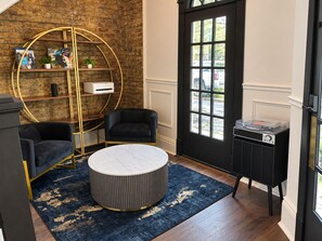 Quarto com parede de tijolos, porta preta, mesa redonda, duas cadeiras pretas, estante e um pequeno armário preto.