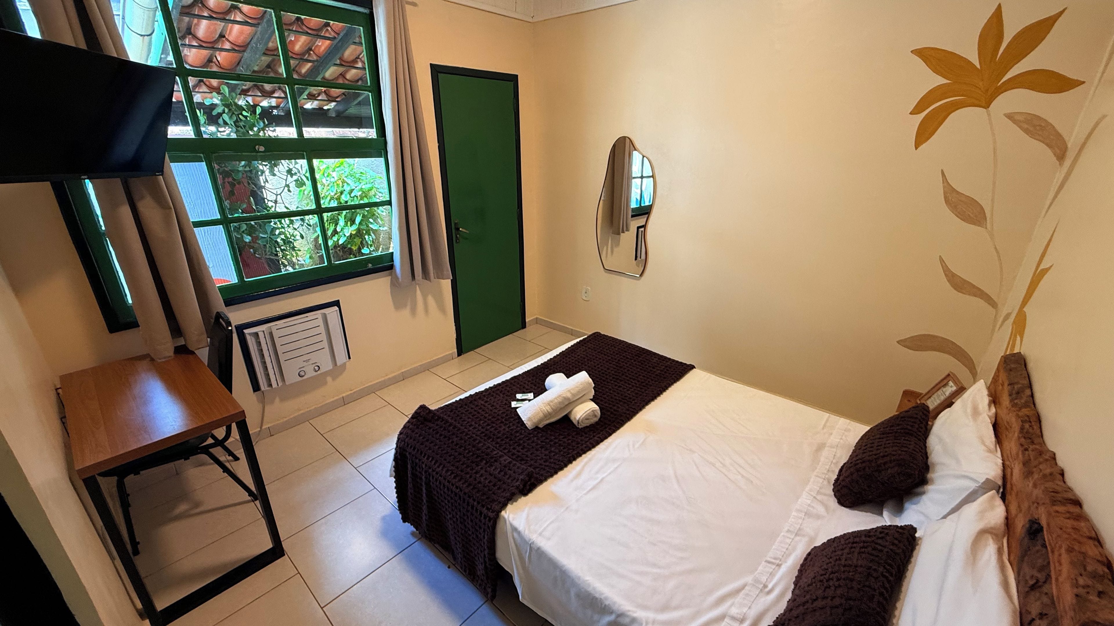 Quarto de hotel com cama, uma mesinha, janela vista para área verde, porta e espelho na parede.