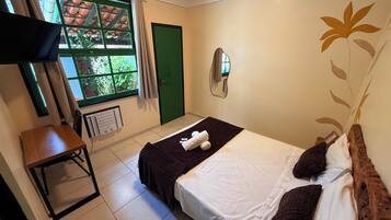 Quarto de hotel com cama, uma mesinha, janela vista para área verde, porta e espelho na parede.