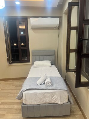 Uma cama de solteiro com cabeceira cinza e roupa de cama branca, um aparelho de ar condicionado acima e uma janela vista para o exterior.