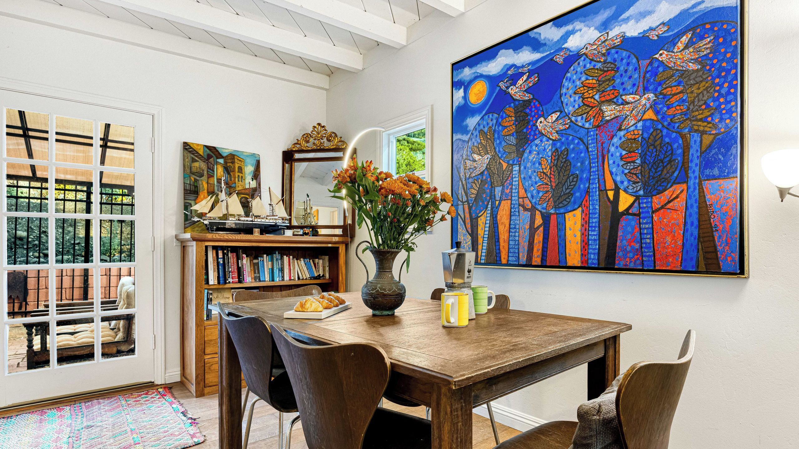 Uma área de refeições com mesa de madeira, cadeiras, um vaso com flores, uma estante de livros e um quadro grande na parede.