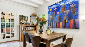 Uma área de refeições com mesa de madeira, cadeiras, um vaso com flores, uma estante de livros e um quadro grande na parede.