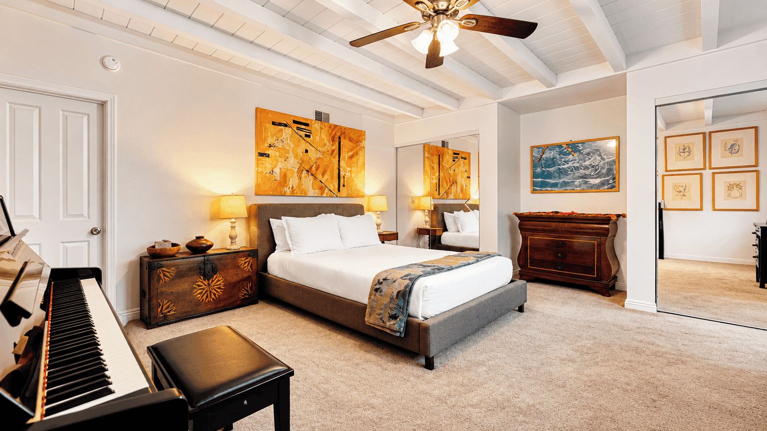 Quarto com piano, cama com cabeceira, ventilador de teto e obras de arte nas paredes.