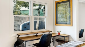 Quarto com escrivaninha, duas cadeiras, janela com vista para uma árvore e um desenho emoldurado na parede.
