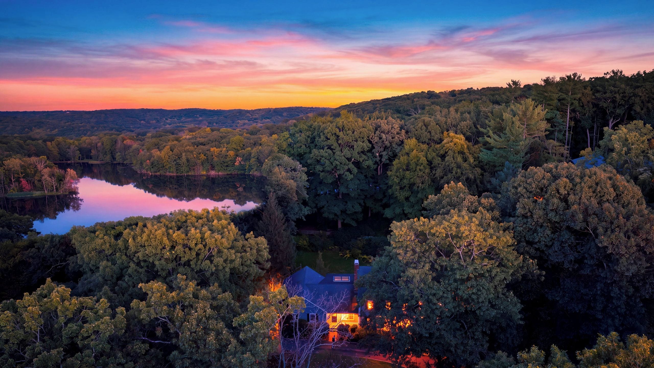Uma propriedade à beira do lago, com uma casa rodeada por uma floresta densa, vista clara do lago e céu ao pôr do sol.