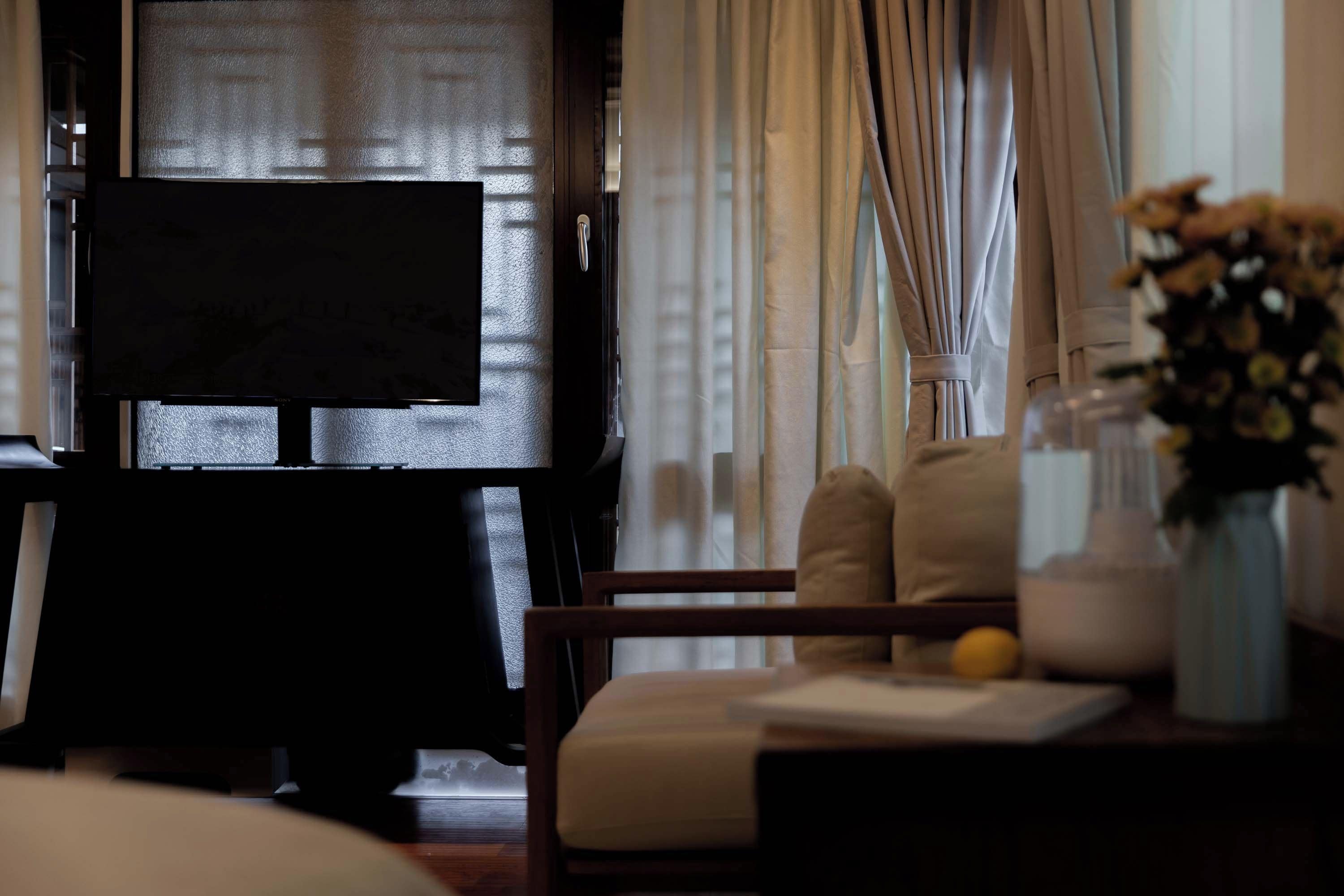 Quarto com televisão, sofá, mesa com um vaso e um livro, e janela com cortinas.