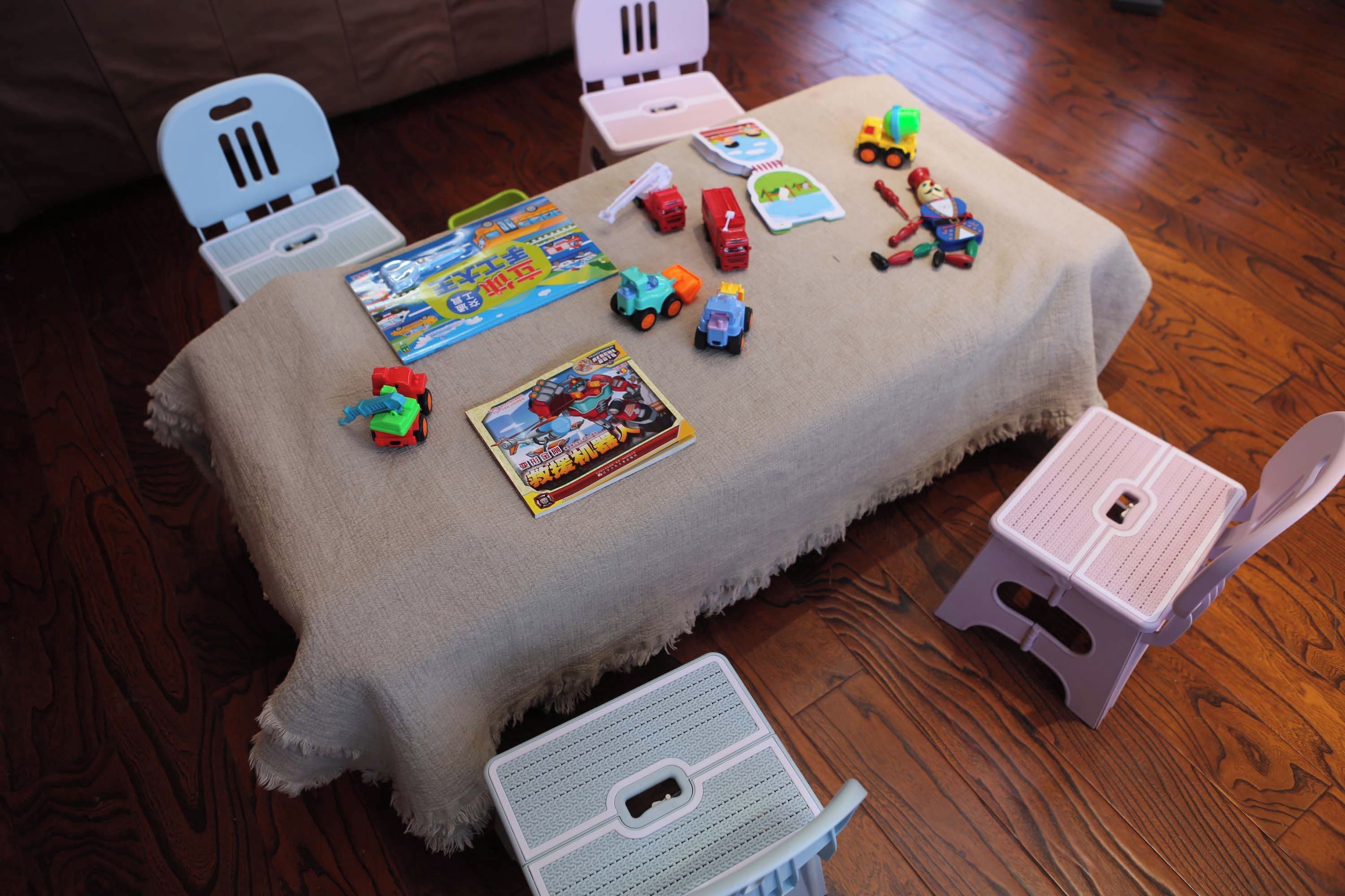 Uma mesa com brinquedos infantis e livros, uma cadeira branca e uma mesinha com um porta-lenços.