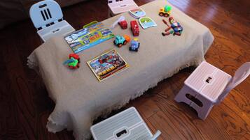Uma mesa com brinquedos infantis e livros, uma cadeira branca e uma mesinha com um porta-lenços.